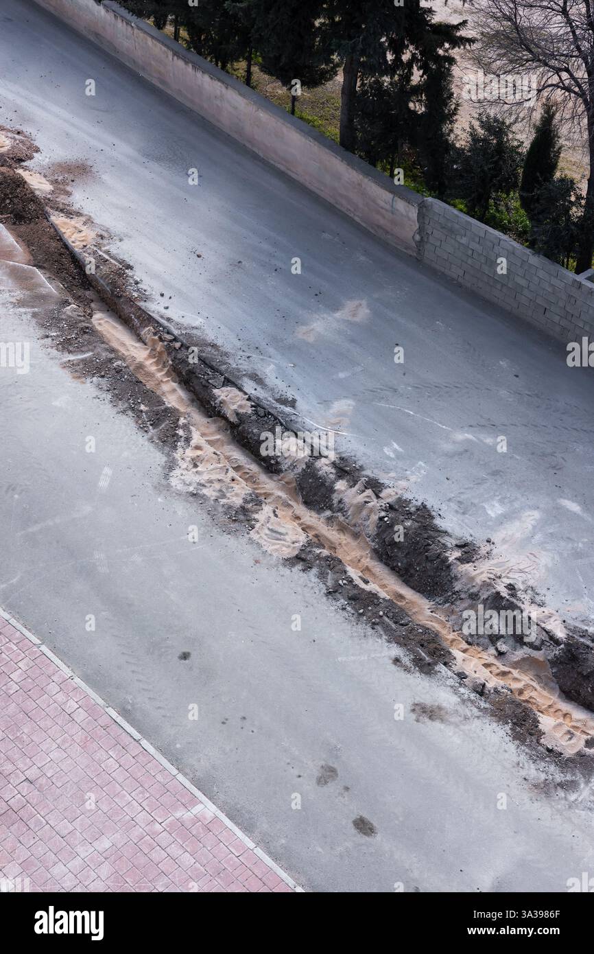 A construction site reveals a freshly dug trench in gray dirt and sand ...