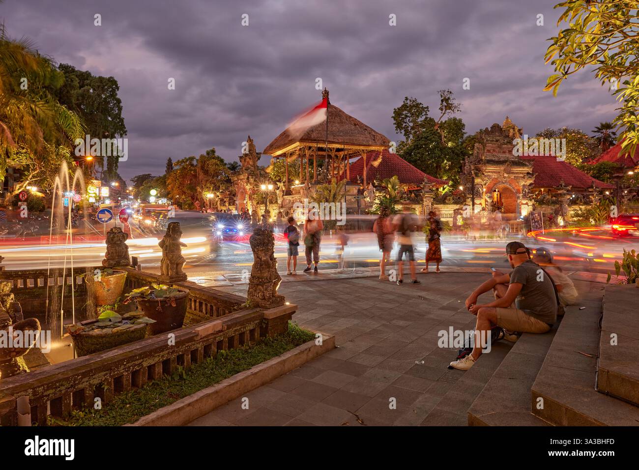 People sit on the steps at Jalan Raya Ubud street illuminated at dusk ...