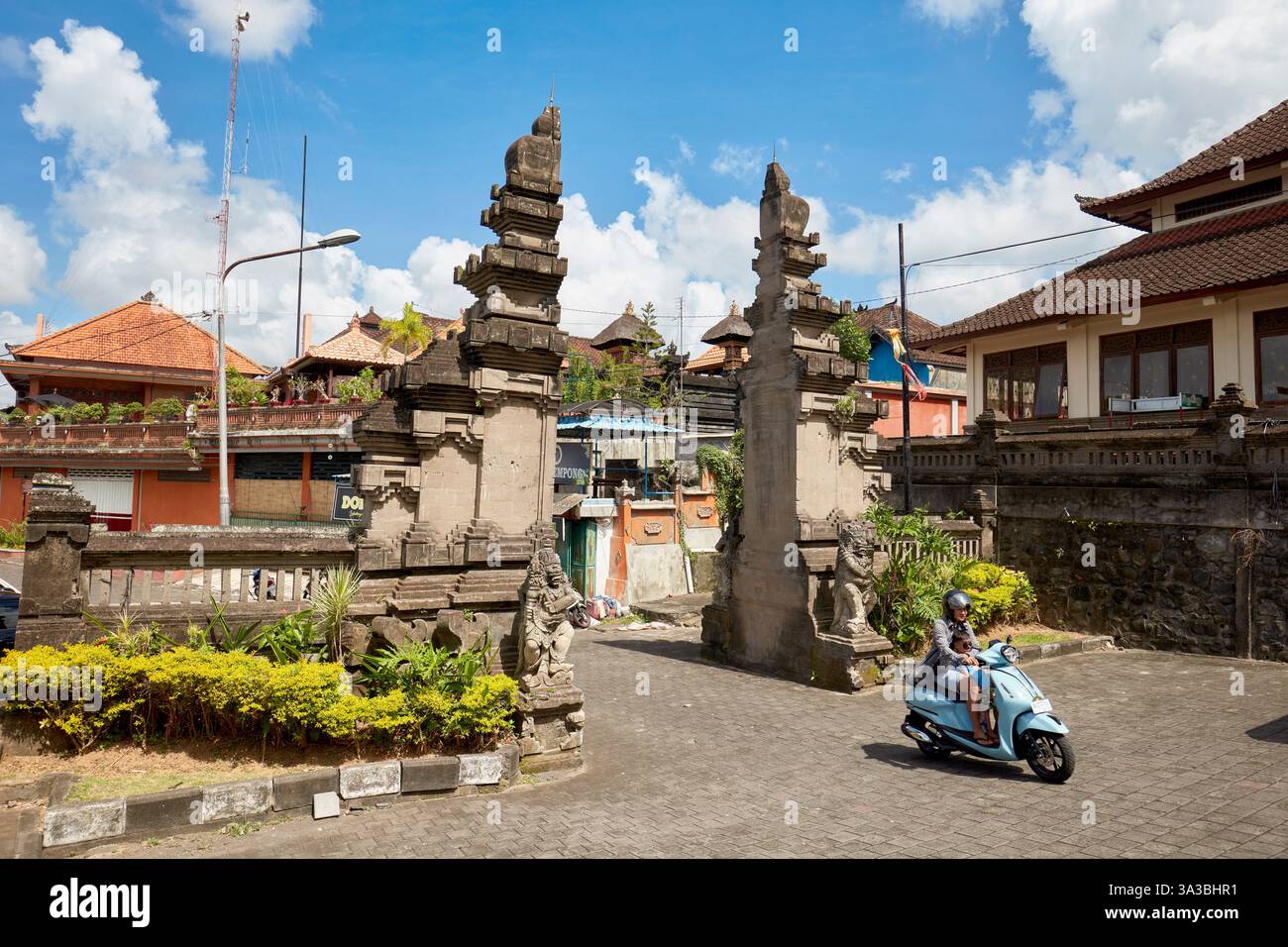 A woman holding her child rides on blue motorbike through candi bentar ...