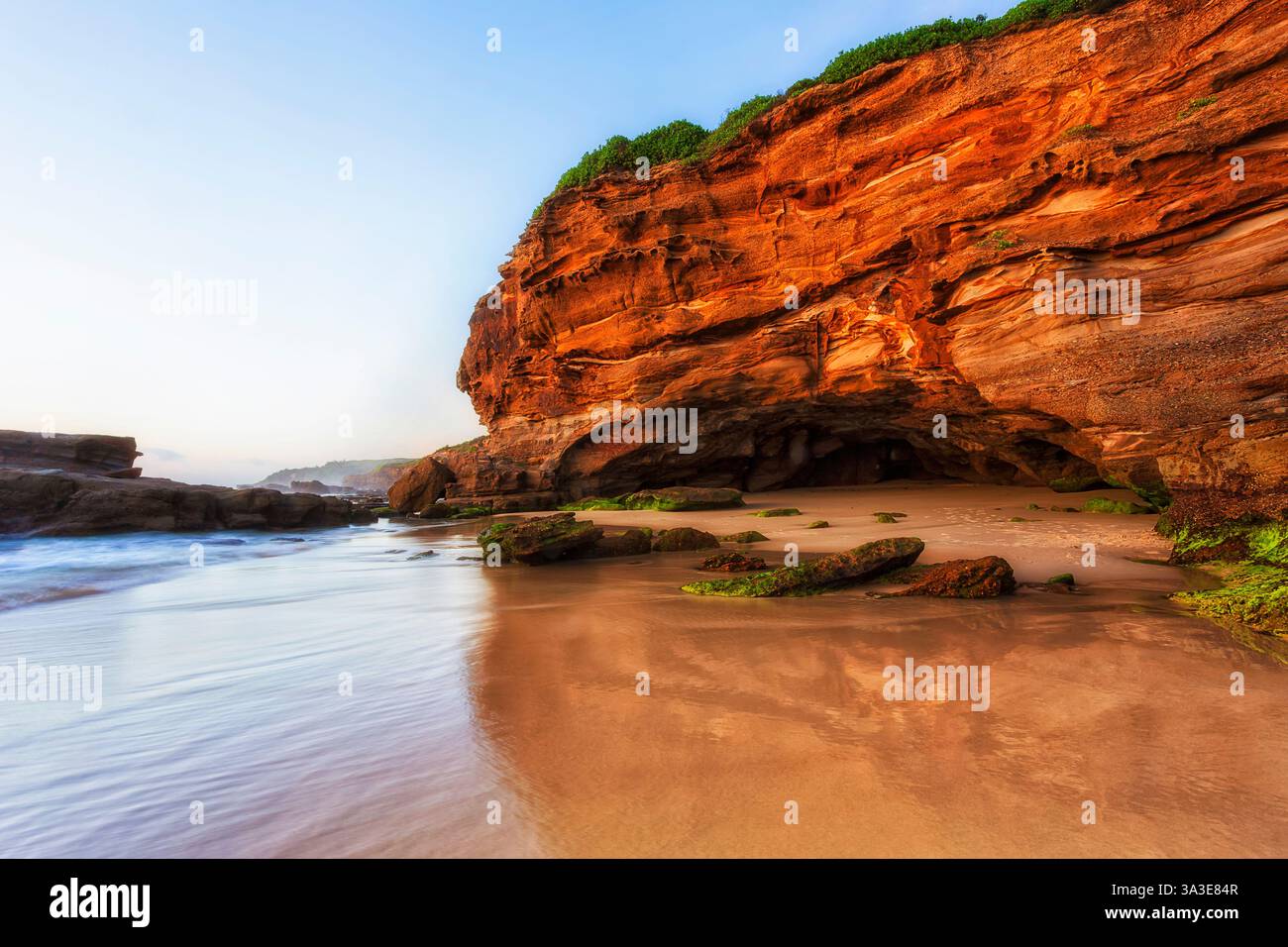 Wet beach sand reflecting scenic sea cave under sandstone cliff on Pacific coast of Australia at ...