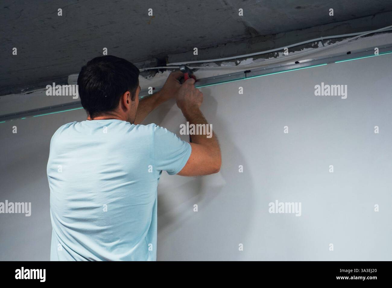 A worker carefully drills into a ceiling, balancing on a ladder. Dust ...