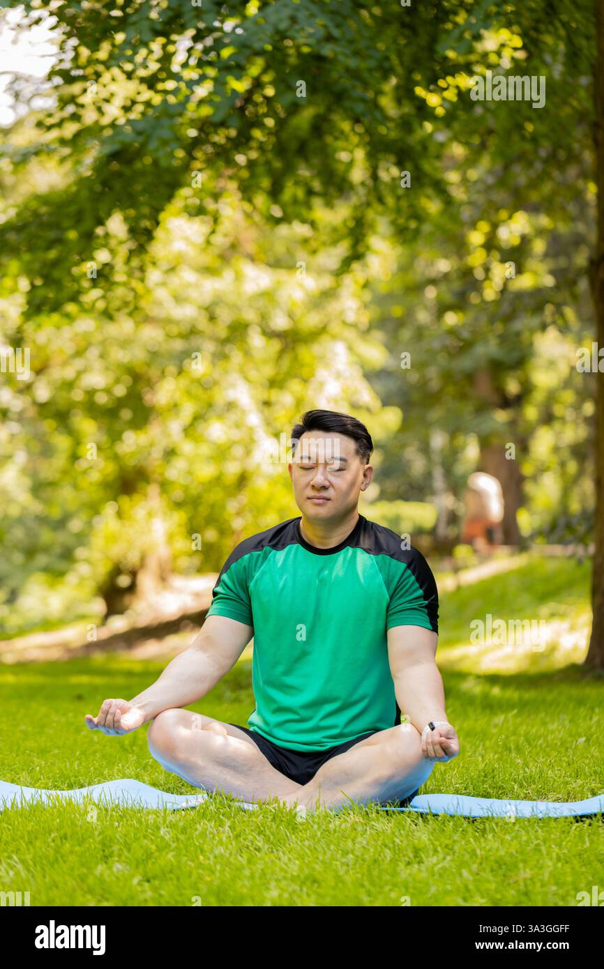 Young Asian man sitting on mat in lotus position, relaxing in city park ...
