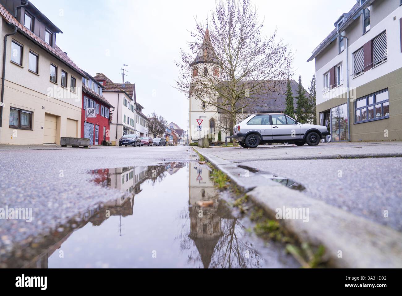Rural road with church tower, puddle creates reflection, cars parked at ...