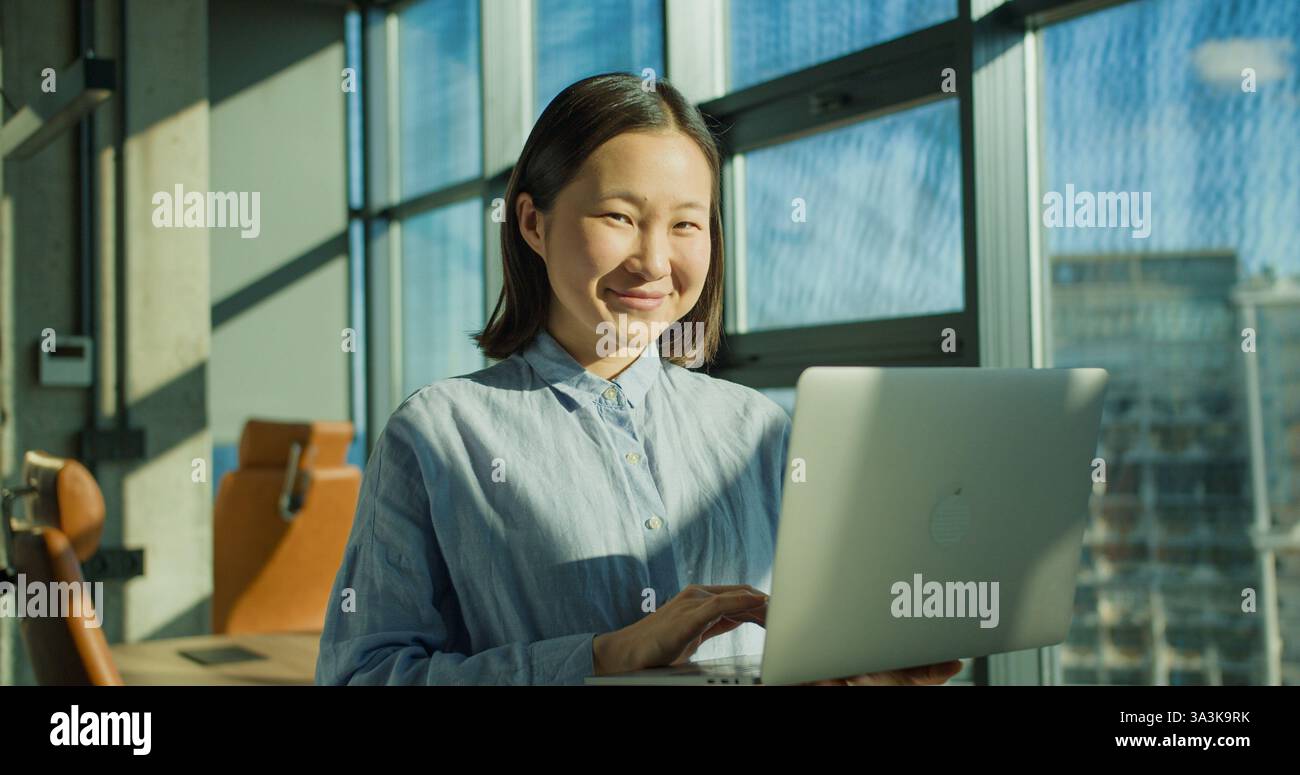 Focused young woman working on a laptop in a modern office, illuminated ...