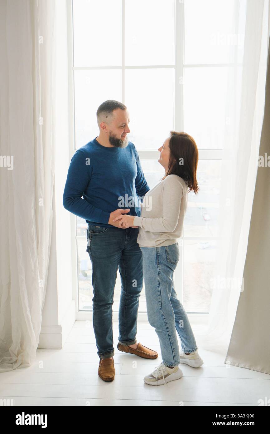 Loving couple at 40s stands by large window in home, enjoying quality ...