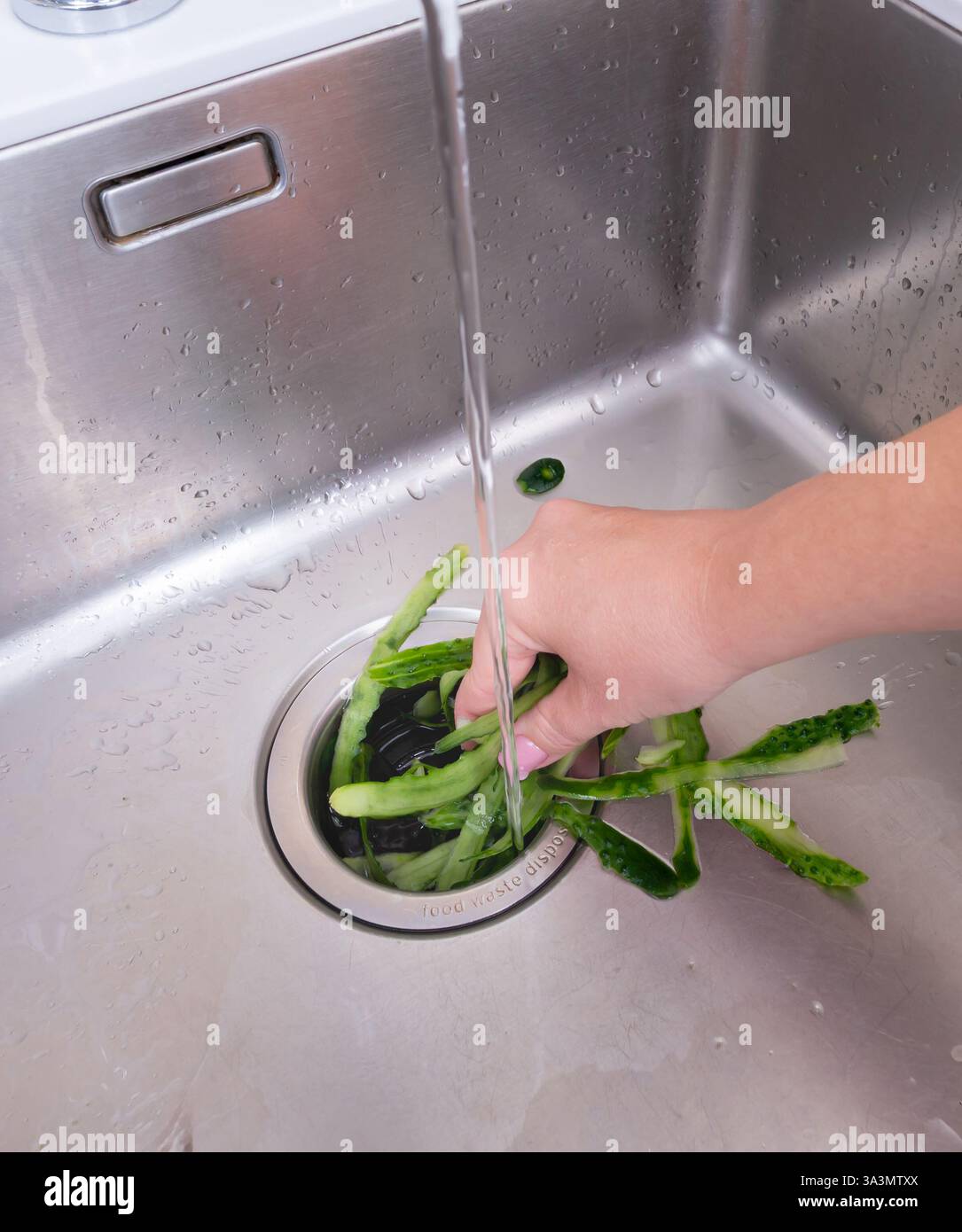Hand disposing of cucumber peels in a stainless steel kitchen sink with ...