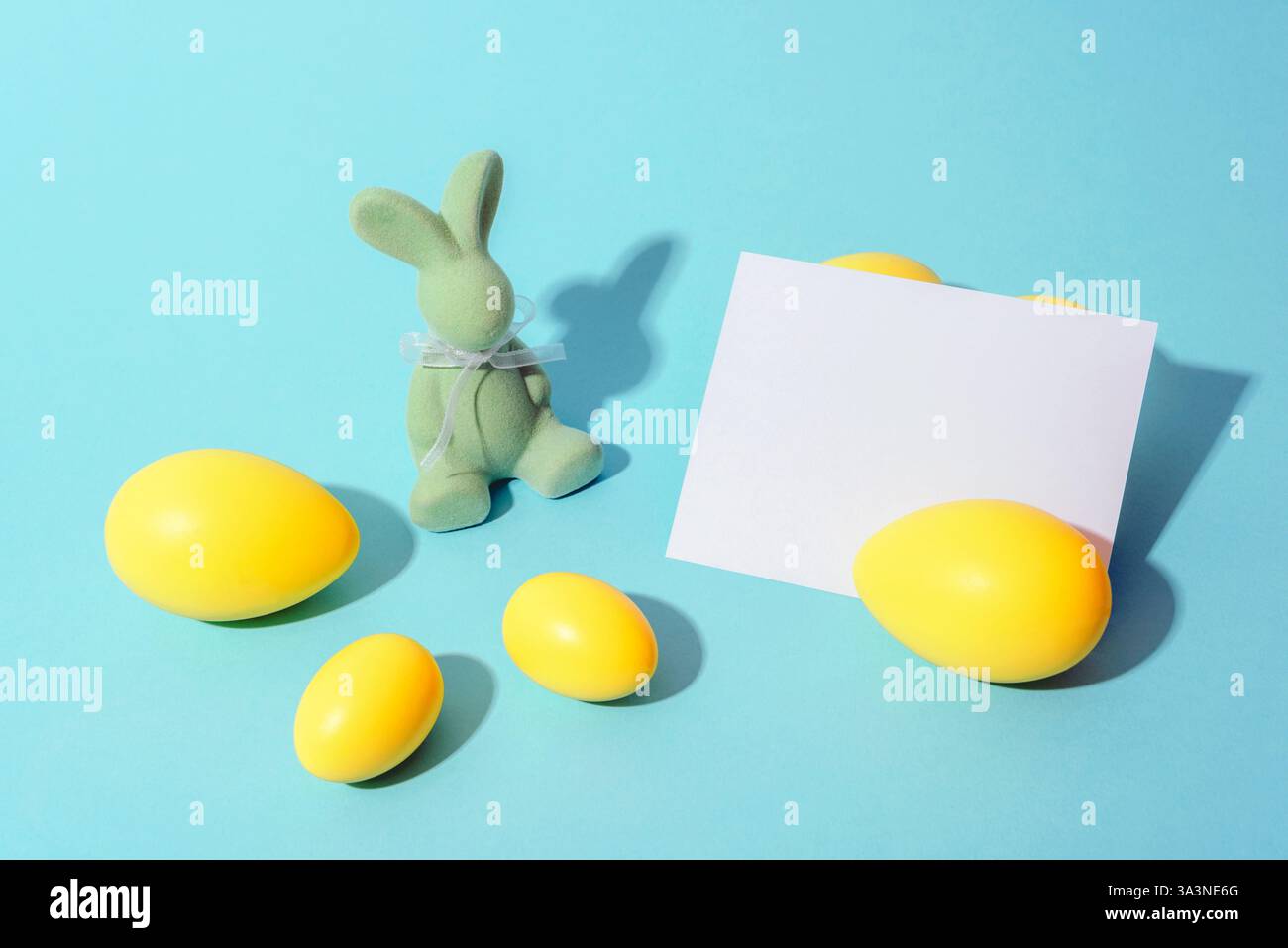 Easter composition with a green velvet bunny, yellow eggs, and a blank white card on a pastel ...