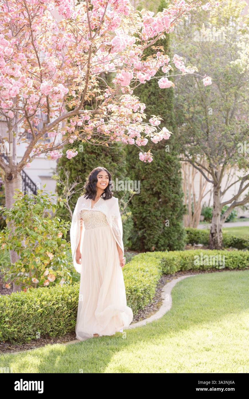 Radiant graduate in a flowing prom dress walks beneath cherry blossoms ...