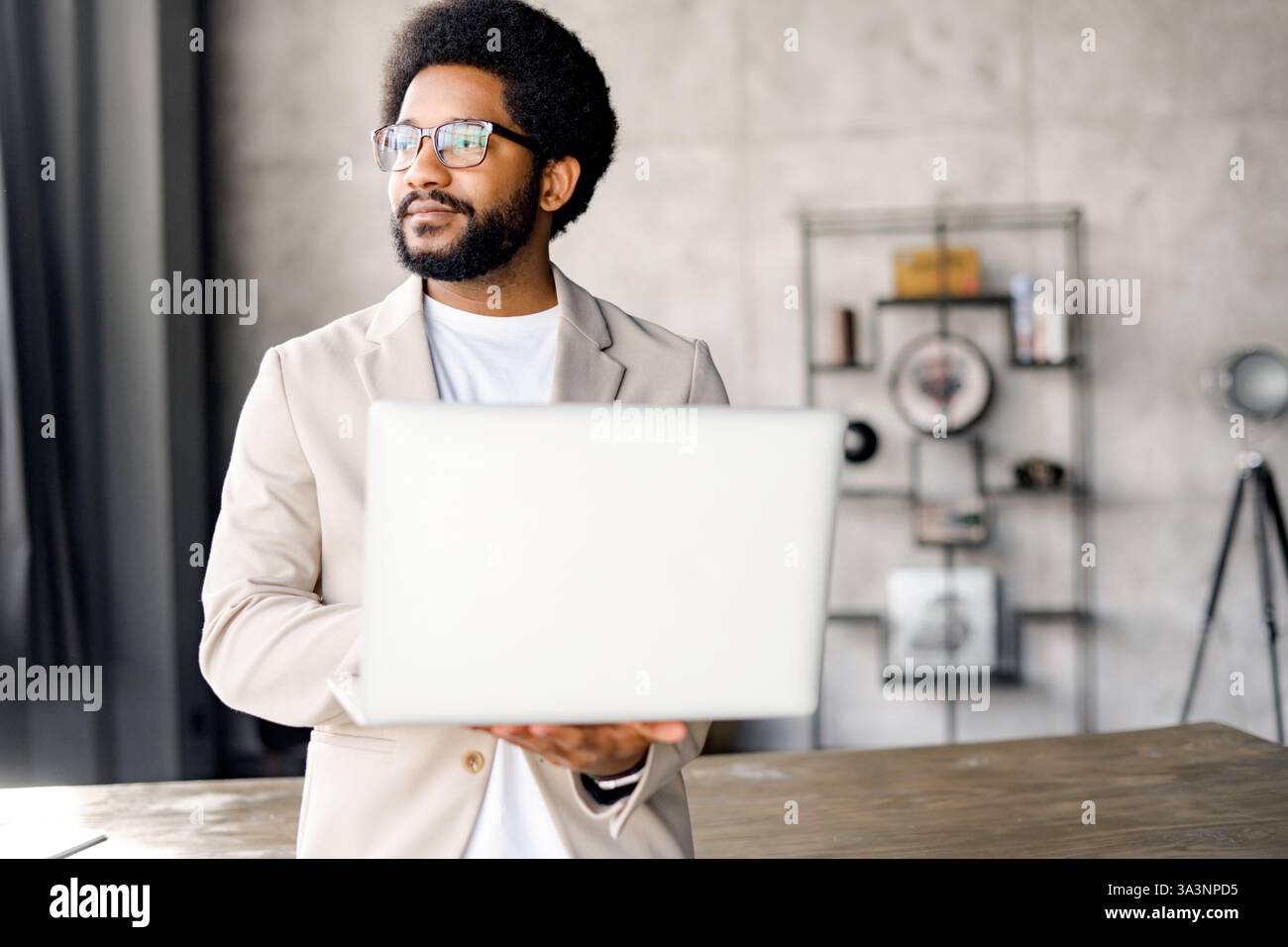 A young Brazilian businessman in suit looks aside thoughtfully, laptop in hand, his demeanor ...