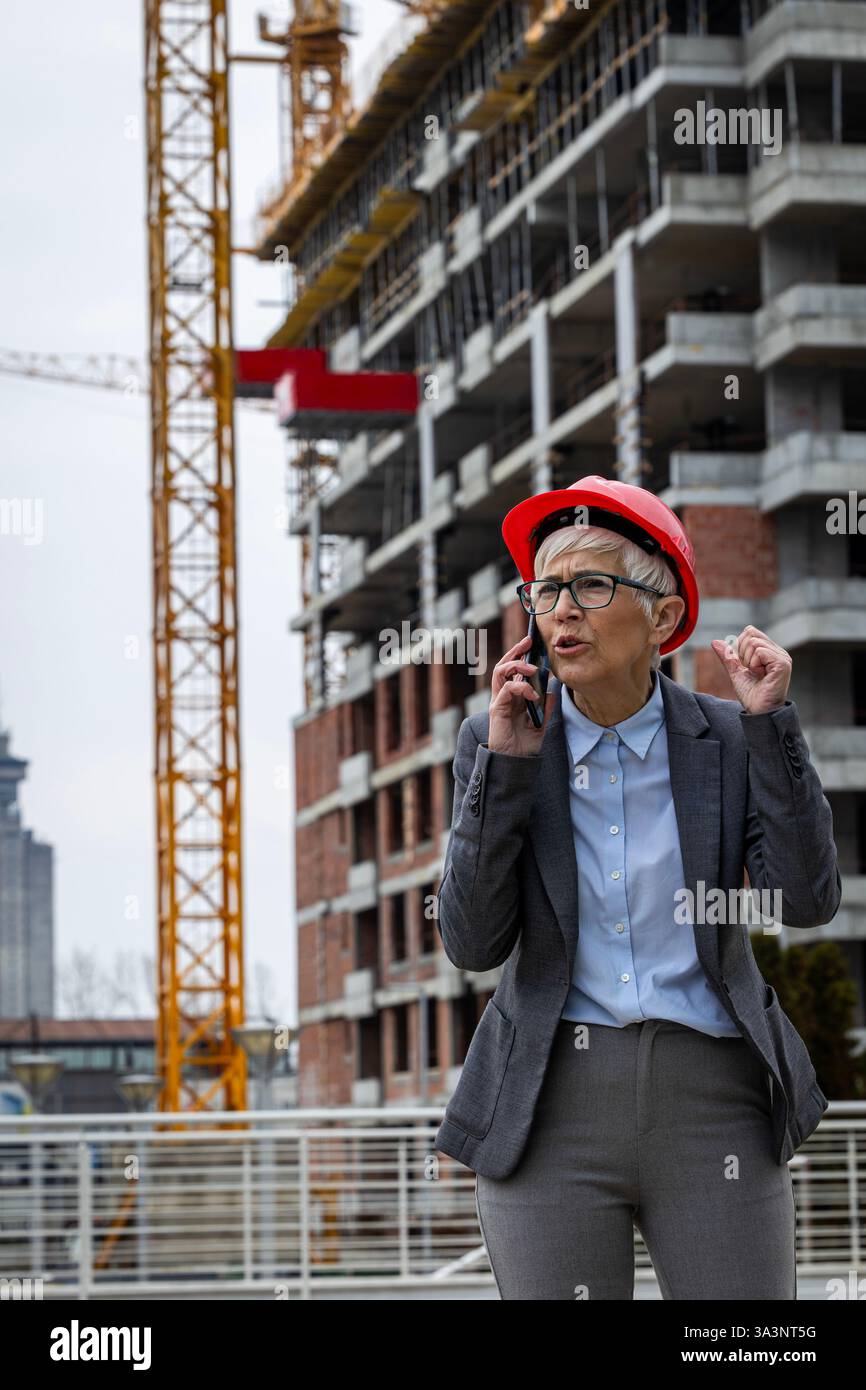 Business woman in gray suit and red hard hat speaking on phone at construction site, managing ...