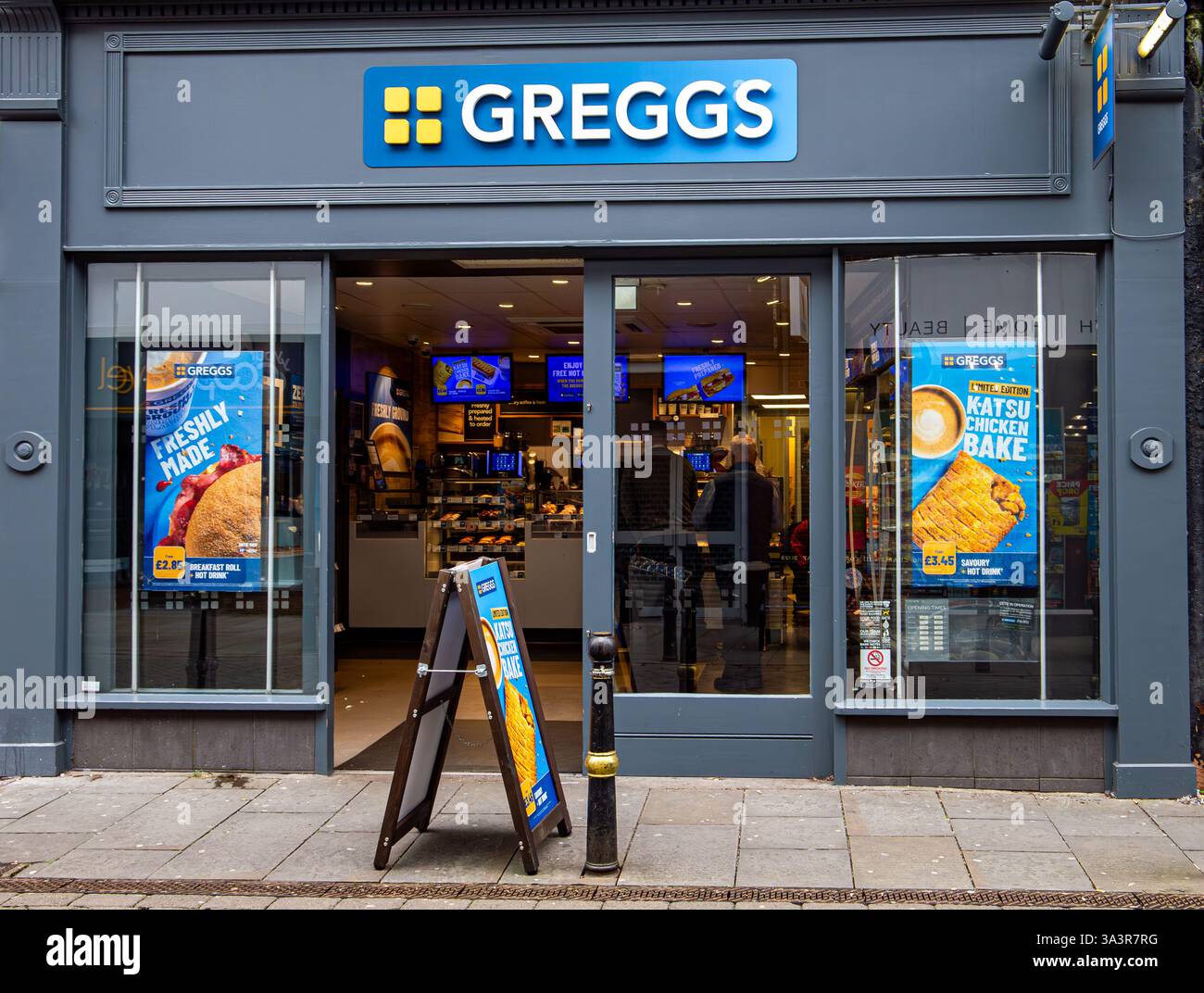 Street view of a Greggs bakery shop entrance with promotional posters ...