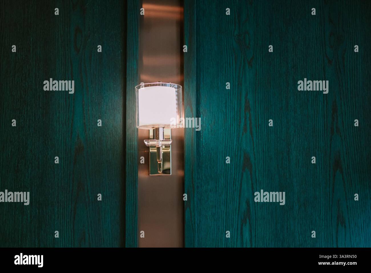A modern wall sconce illuminates a dark teal wood paneled wall. The ...