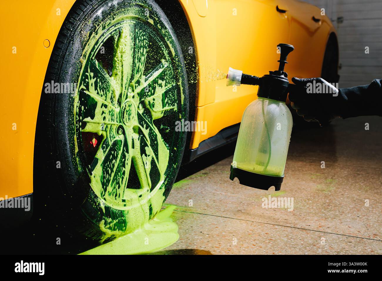 Worker cleans the tire of a yellow car using a spray with soap solution ...