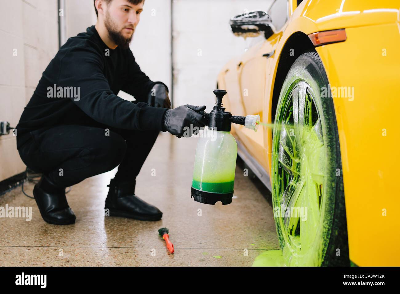 Worker cleans the tire of a yellow car using a spray with soap solution ...