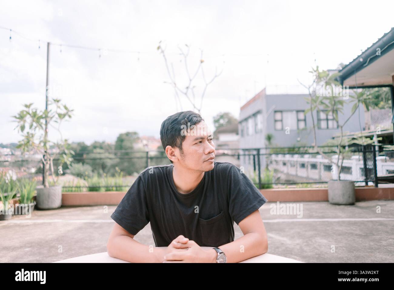 An asian man sits at an outdoor table on a rooftop terrace, gazing ...
