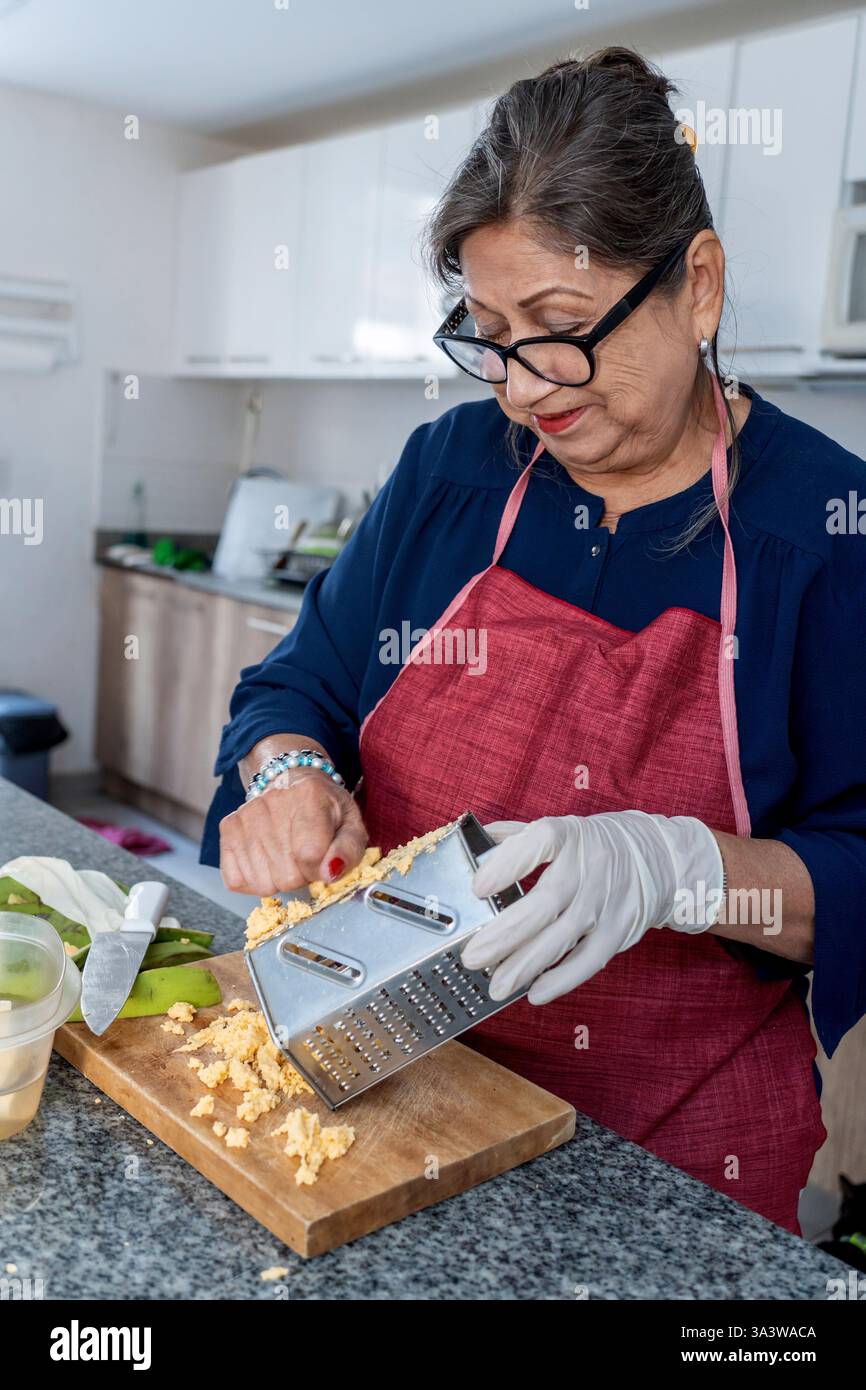 Senior latin woman wearing gloves grating green banana in a modern ...
