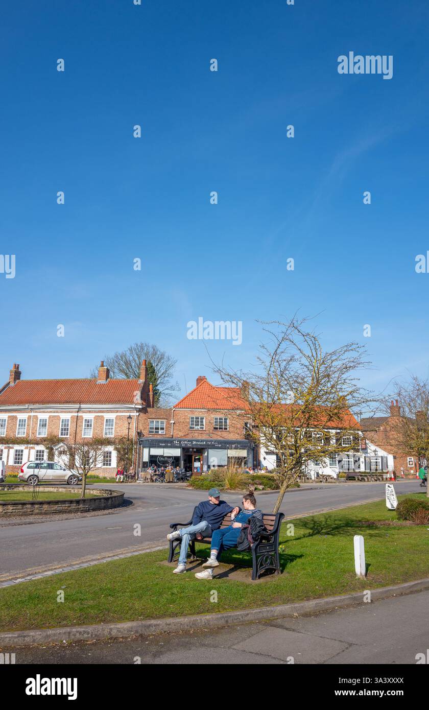 Couple talking sat on a wooden bench on a grass verge in the market ...