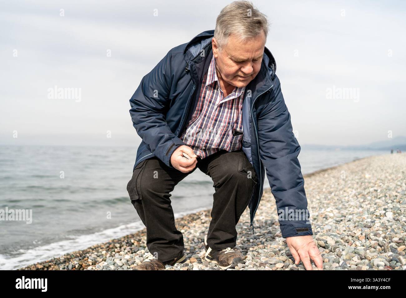 Older male bending down to collect rocks on a pebble beach, engaging ...