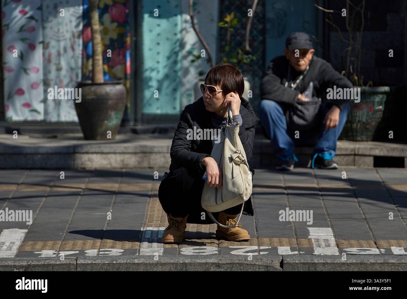 A woman squats as she and another commuter wait for buses at a bus ...