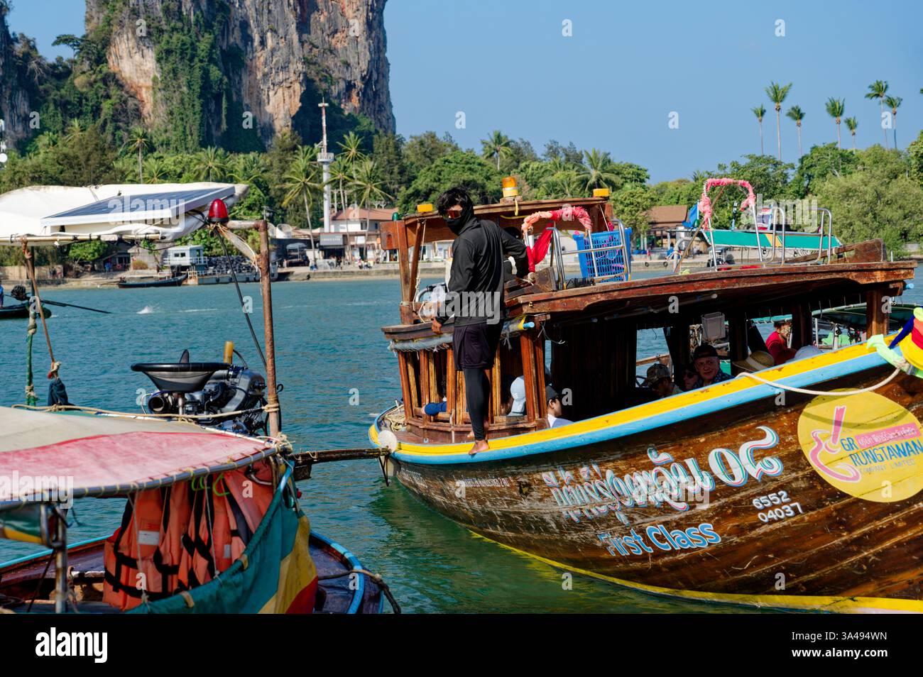 A boat worker balances barefoot on the edge of a wooden long-tail boat ...