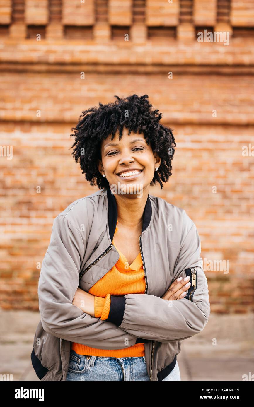 Portrait of a cheerful african american woman standing with crossed arms against a brick wall ...