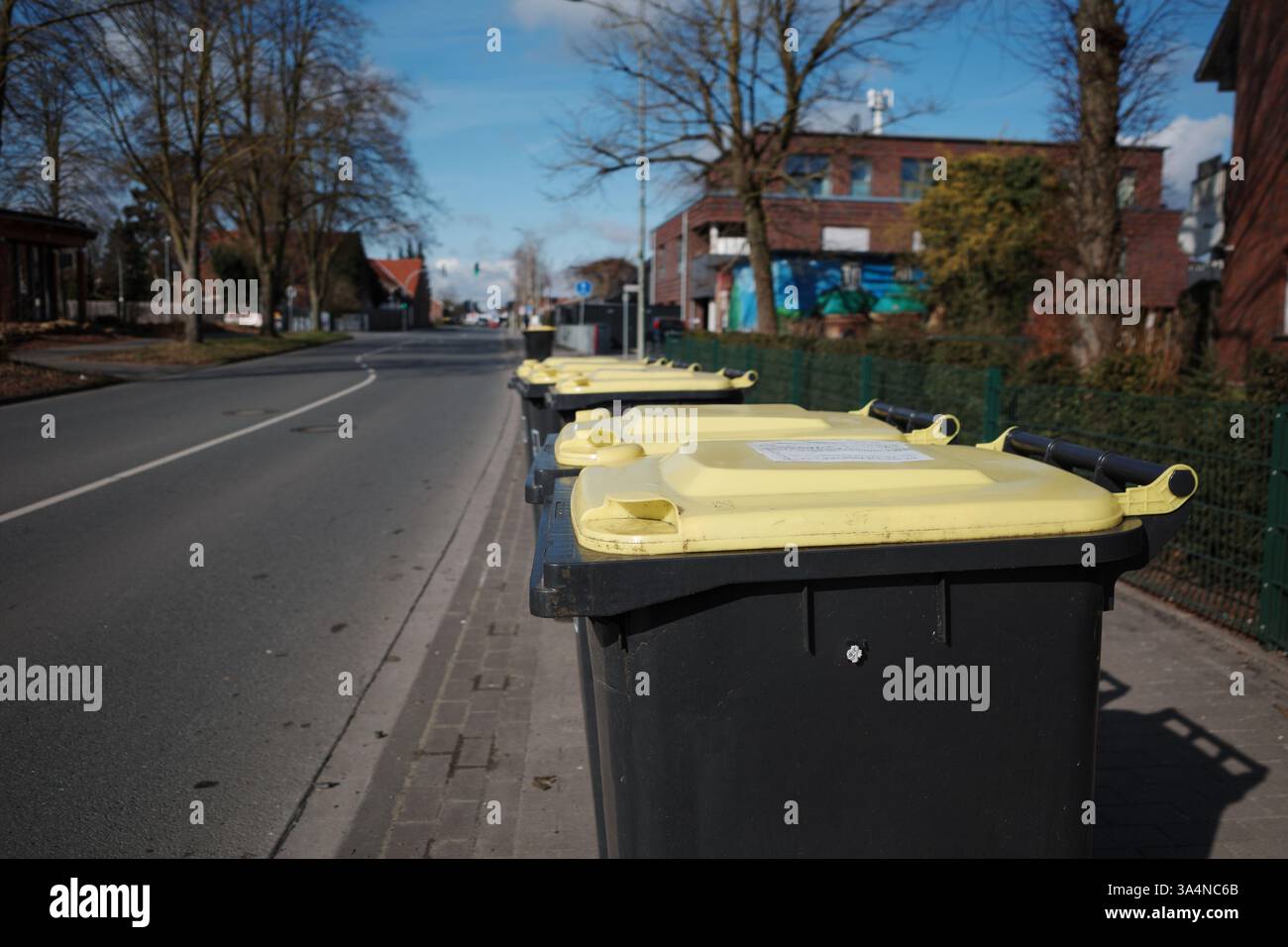 A row of black garbage bins with yellow lids lined up along a sidewalk ...