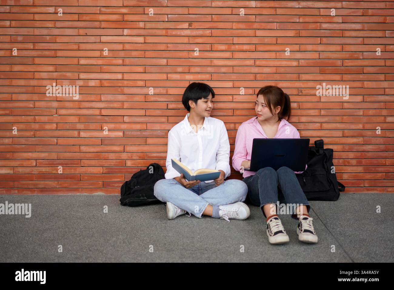 two asian college students seated on the ground, leaning against a ...