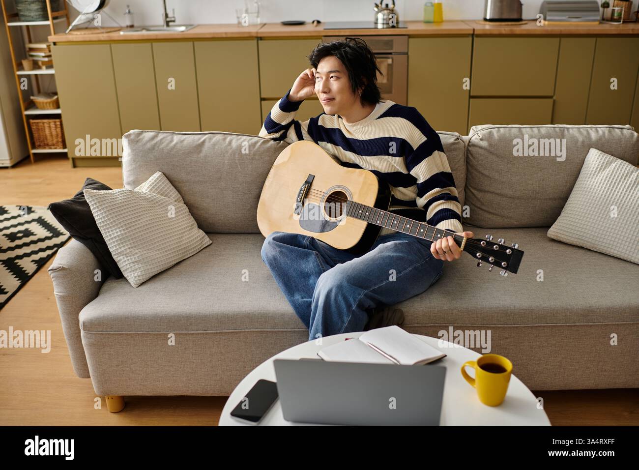 A young man sits relaxed on a couch, practicing his acoustic guitar ...