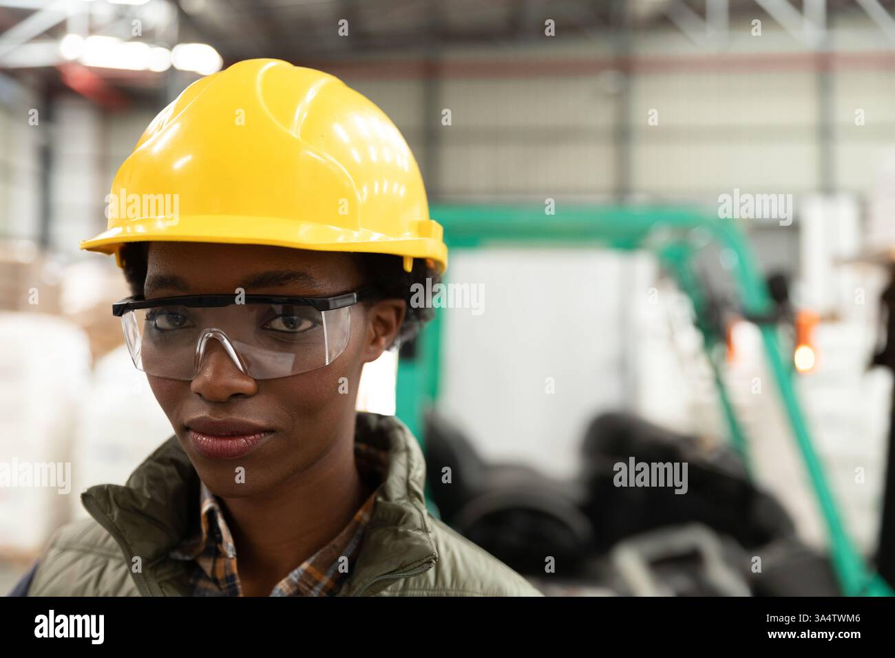 Wearing safety helmet and goggles, warehouse worker confidently standing by forklift, copy space ...
