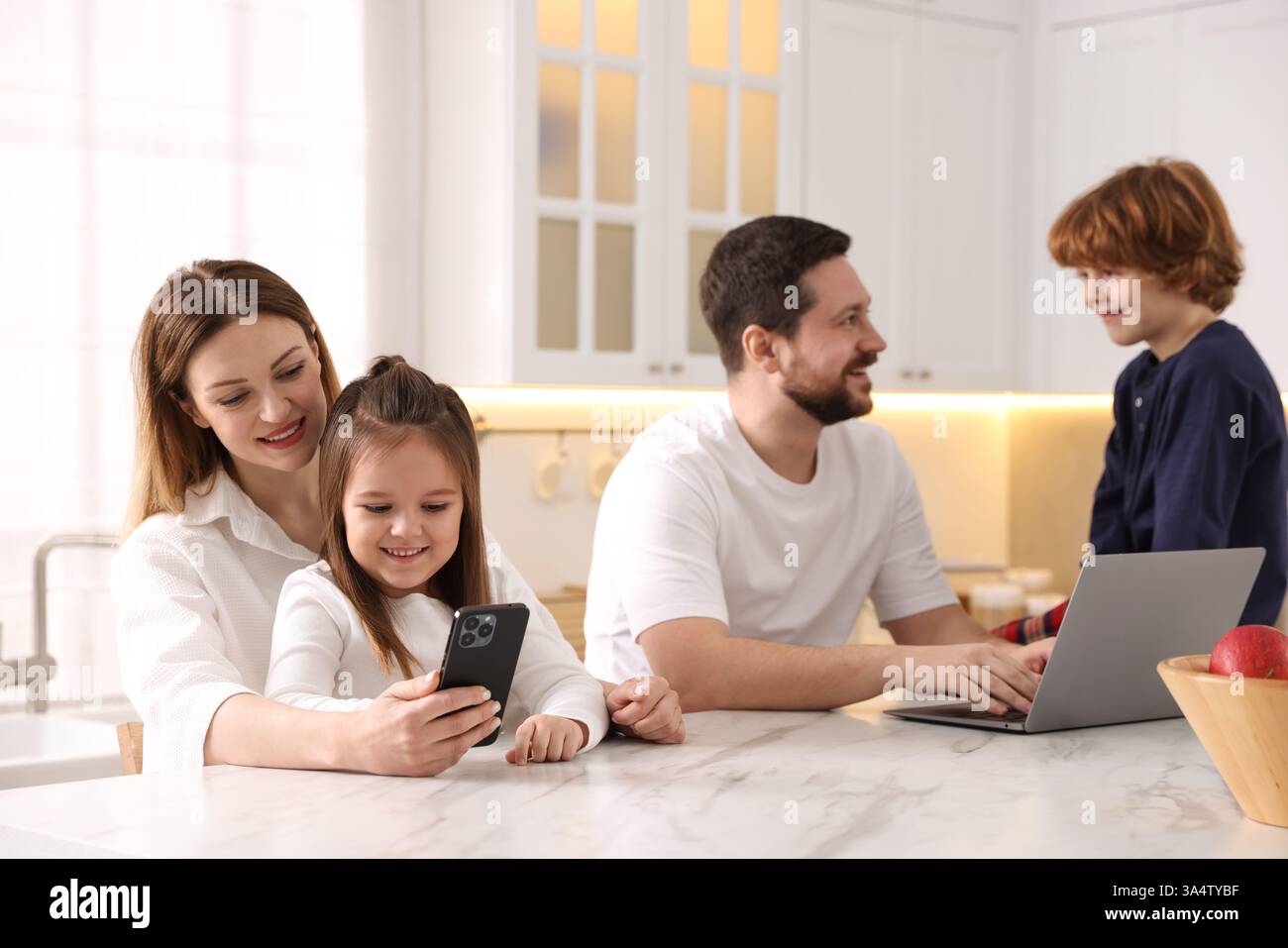 Happy family wearing stylish pajamas looking at devices at white marble ...