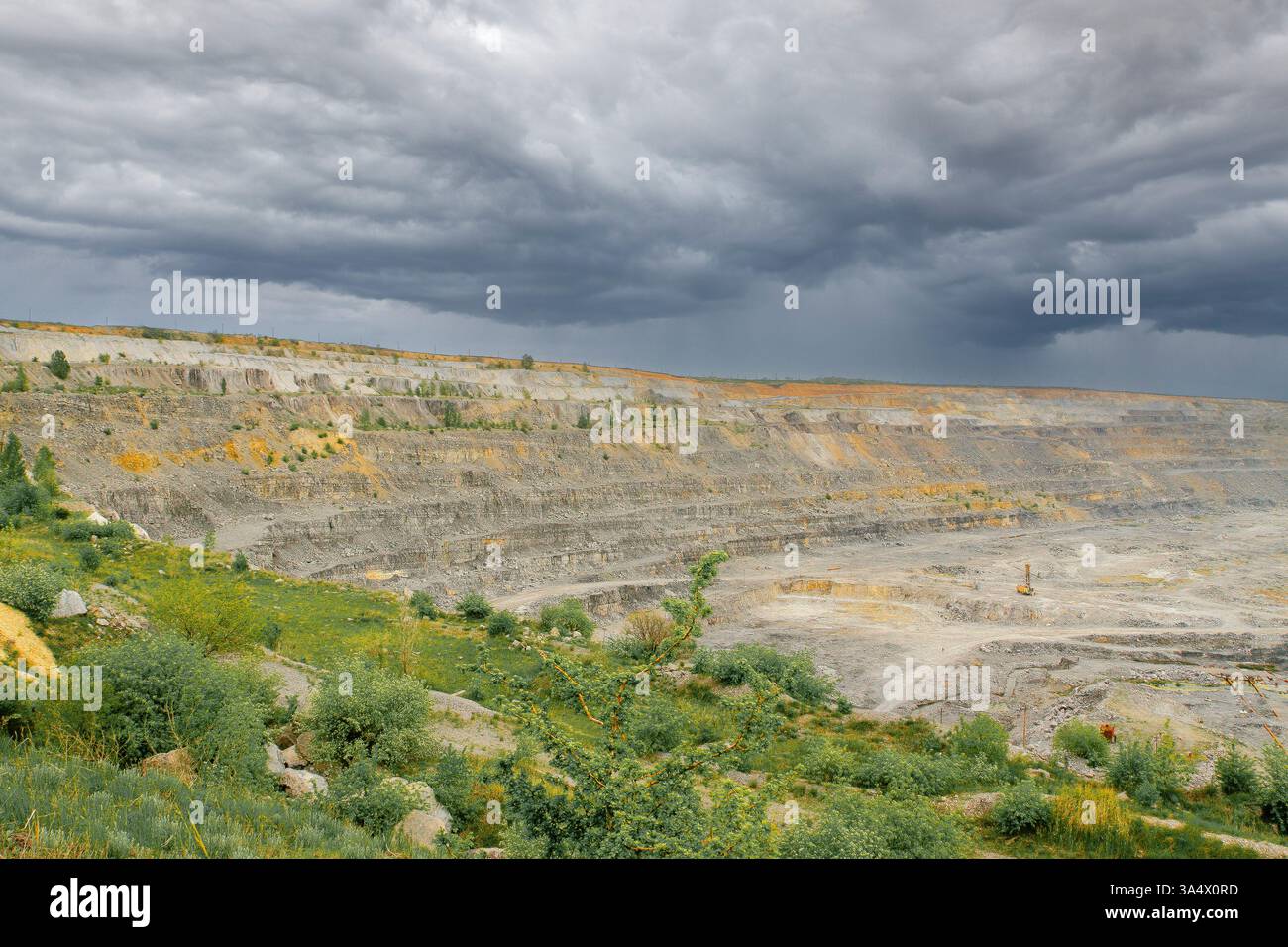 A vast open pit mine under dark storm clouds with dirt roads ...