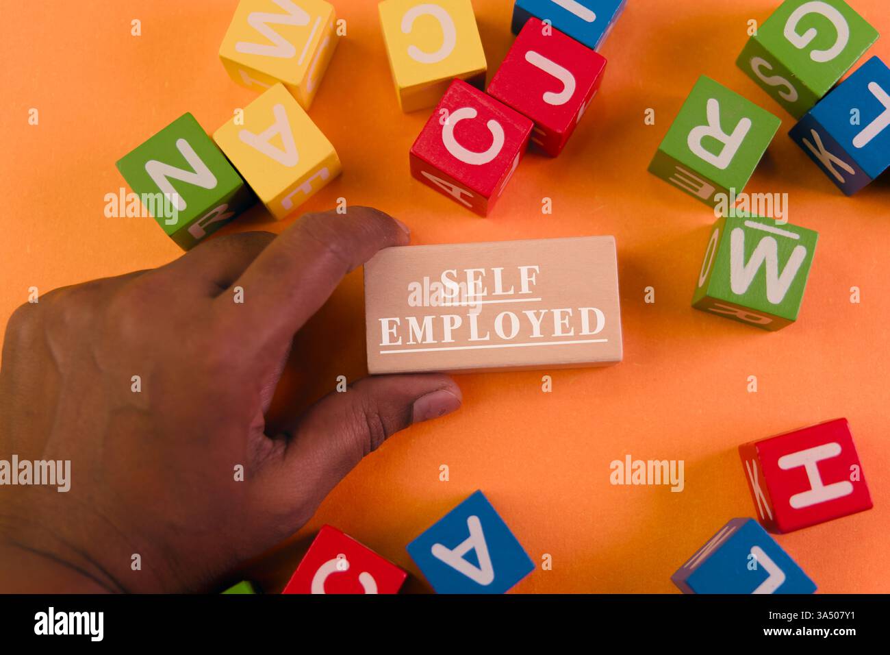 A person’s hand holds a block marked self employed among vibrant letter ...