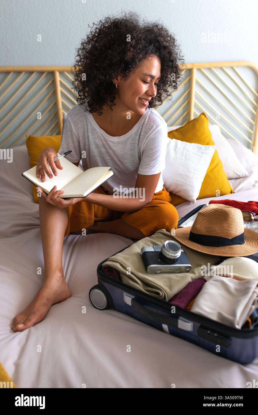 Young woman sitting on bed checking packing list before summer holidays ...