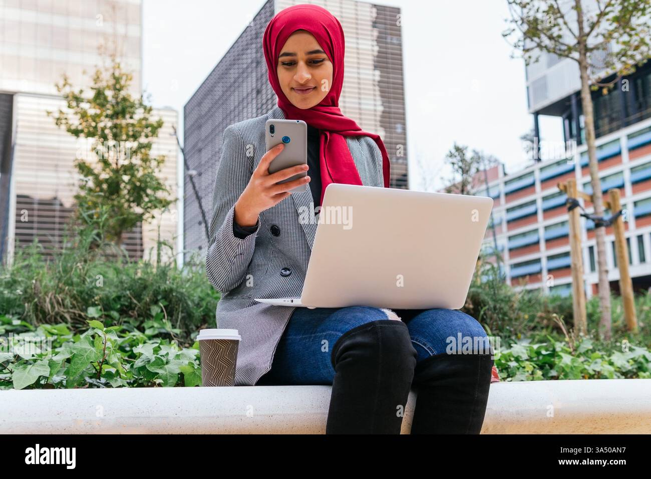 Arab woman in red hijab sitting on bench with laptop using smartphone ...