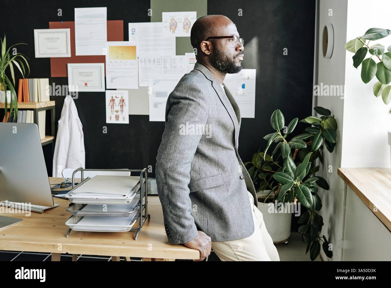 African general practitioner leaning on his office desk and looking at ...