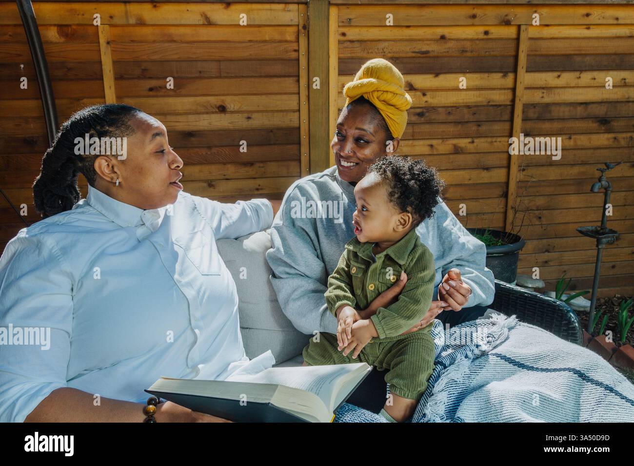 Black non-binary person with dreadlocks reading book with his wife while sitting on chair with ...