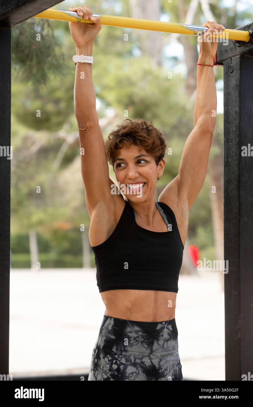 Smiling Hispanic woman performing a bar hang exercise in a park during ...