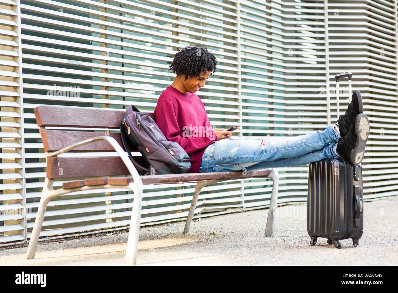 Happy male traveler using mobile phone while sitting on bench and ...