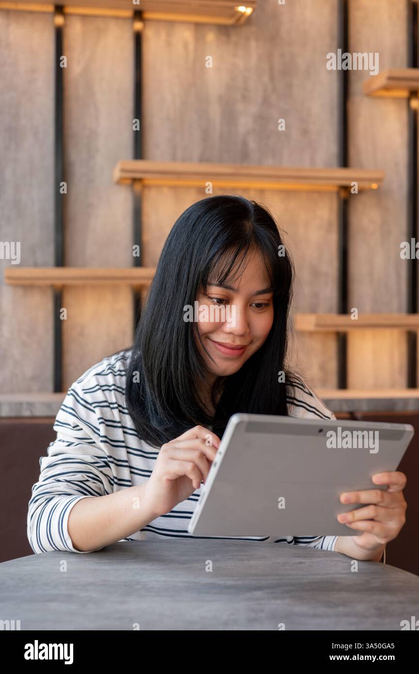 Smiling Asian woman holding and browsing on her digital tablet while sitting inside a coffee ...
