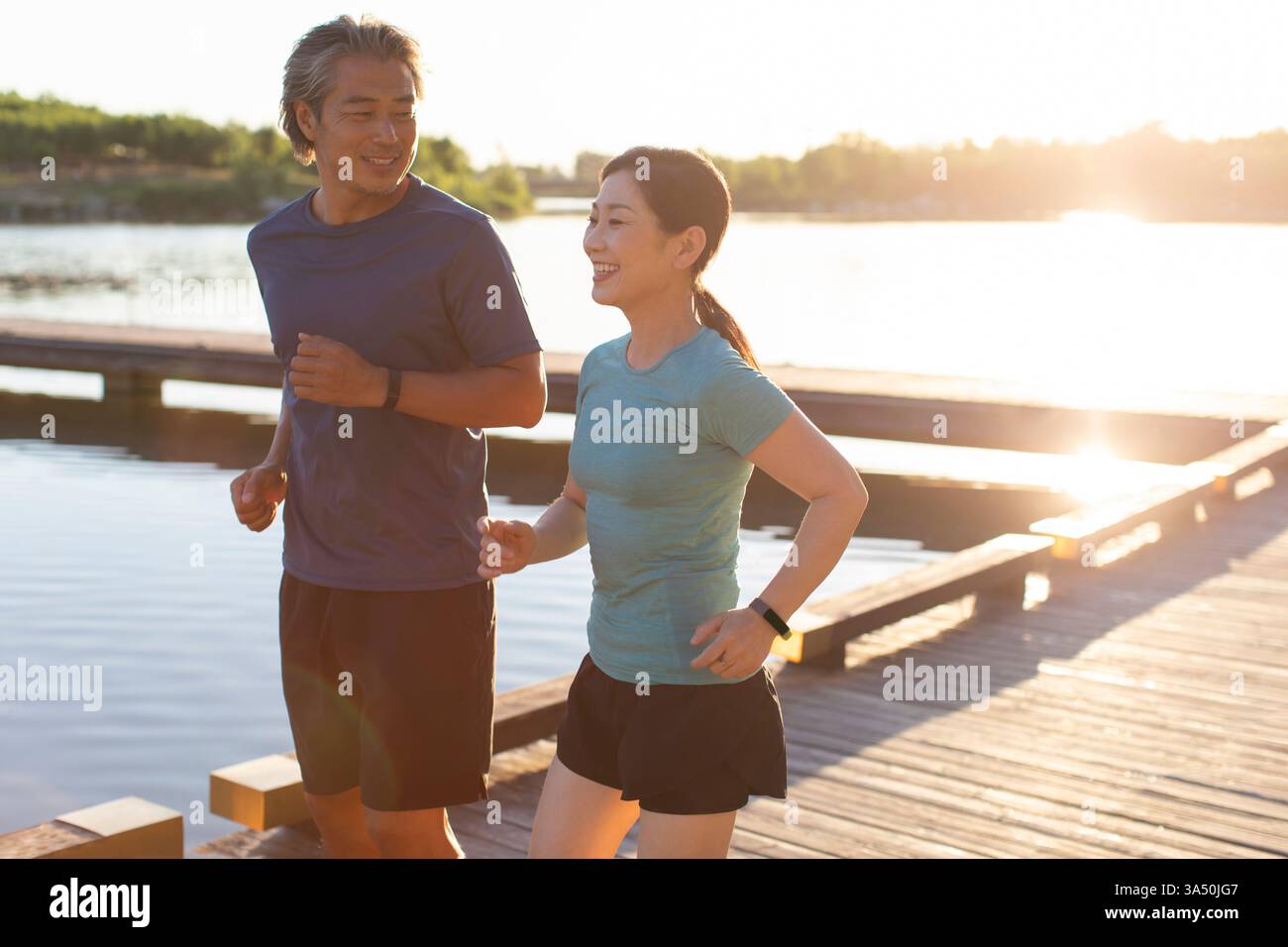 Smiling Chinese couple in their 50s jog together through a lakeside ...