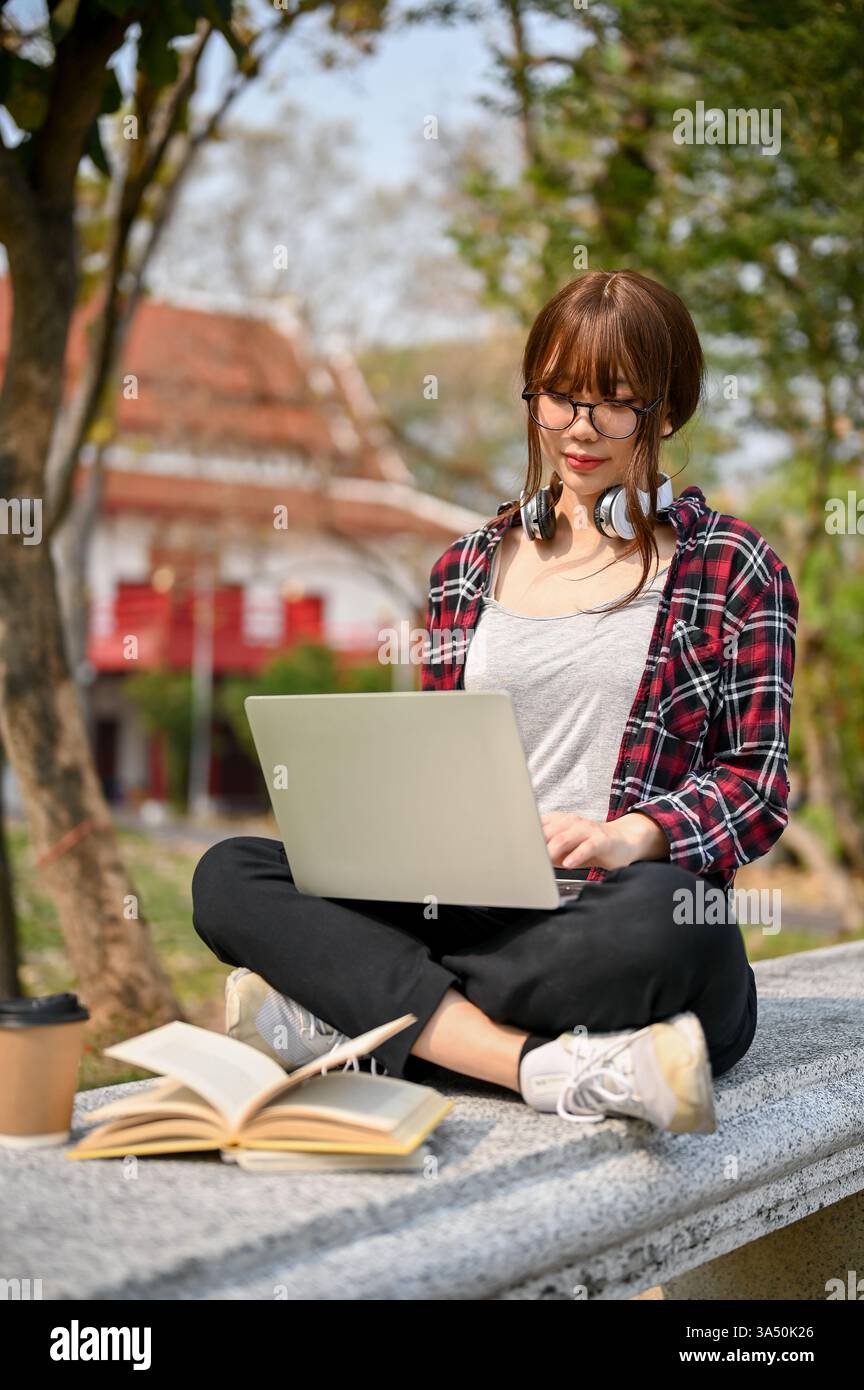 Asian female college student wearing eyeglasses and headphones around neck using laptop studying ...