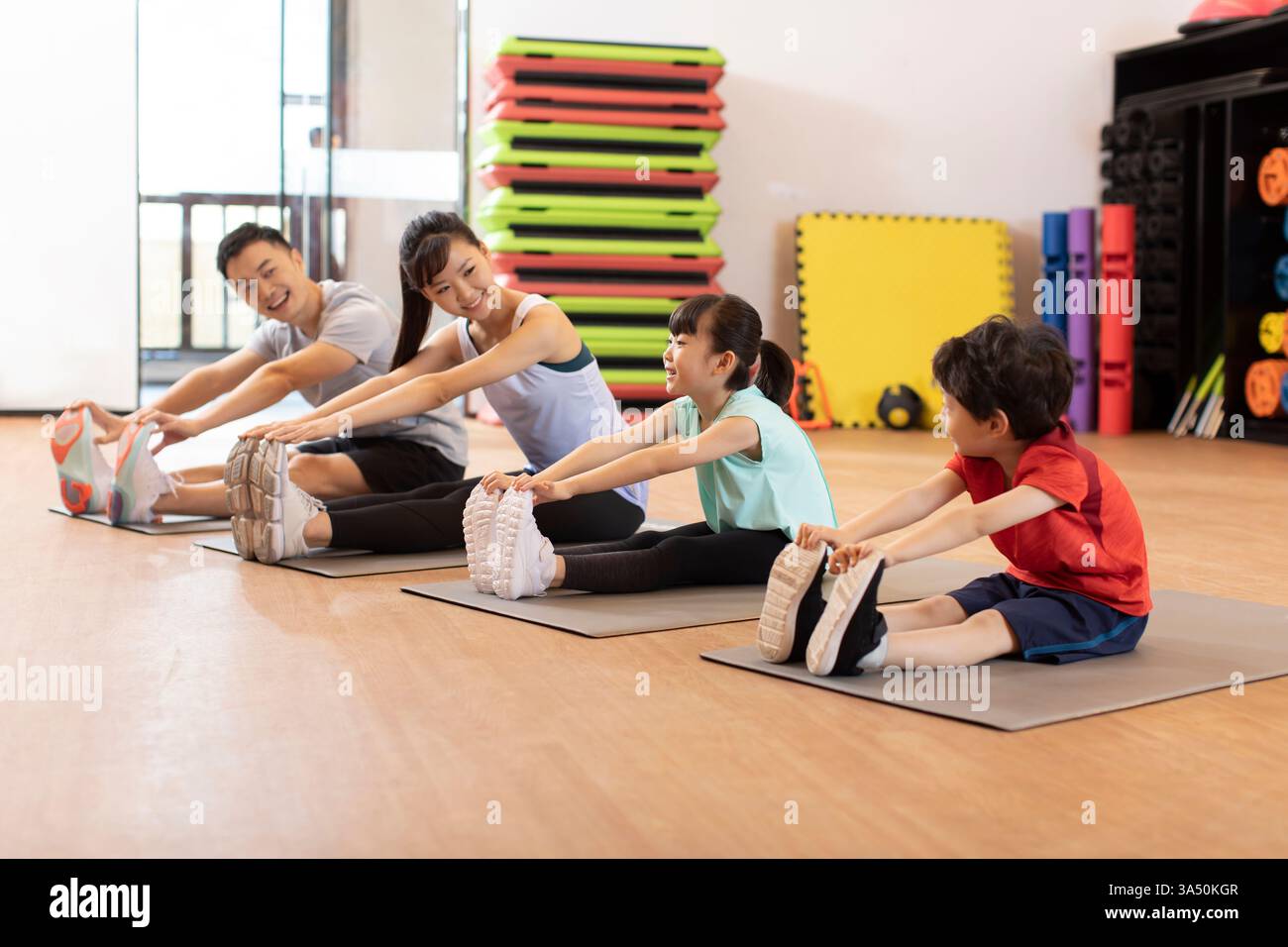Smiling Chinese family doing stretching exercises sitting on mat in gym ...