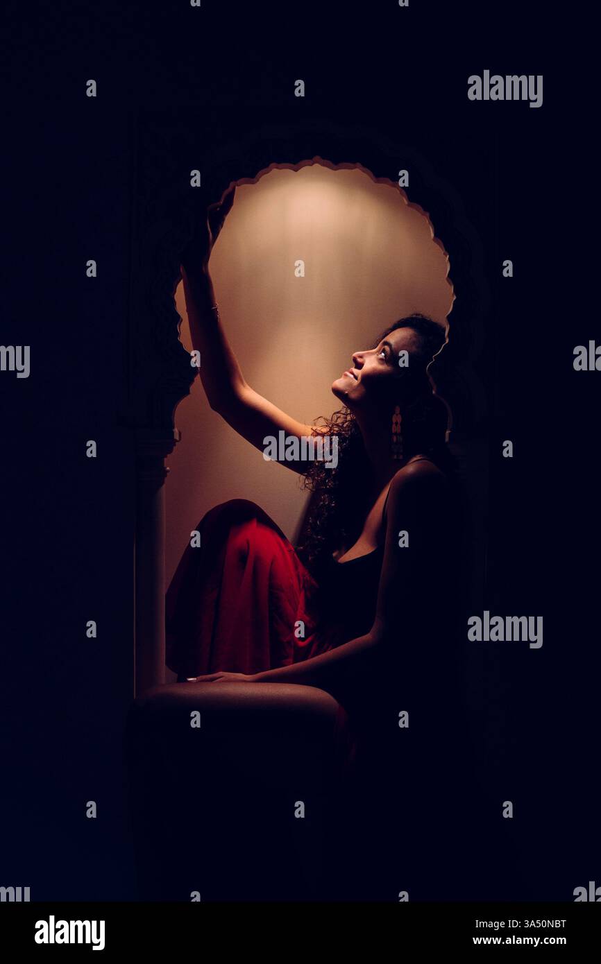 Smiling Arab woman with raised arm looking up sitting on windowsill of ...