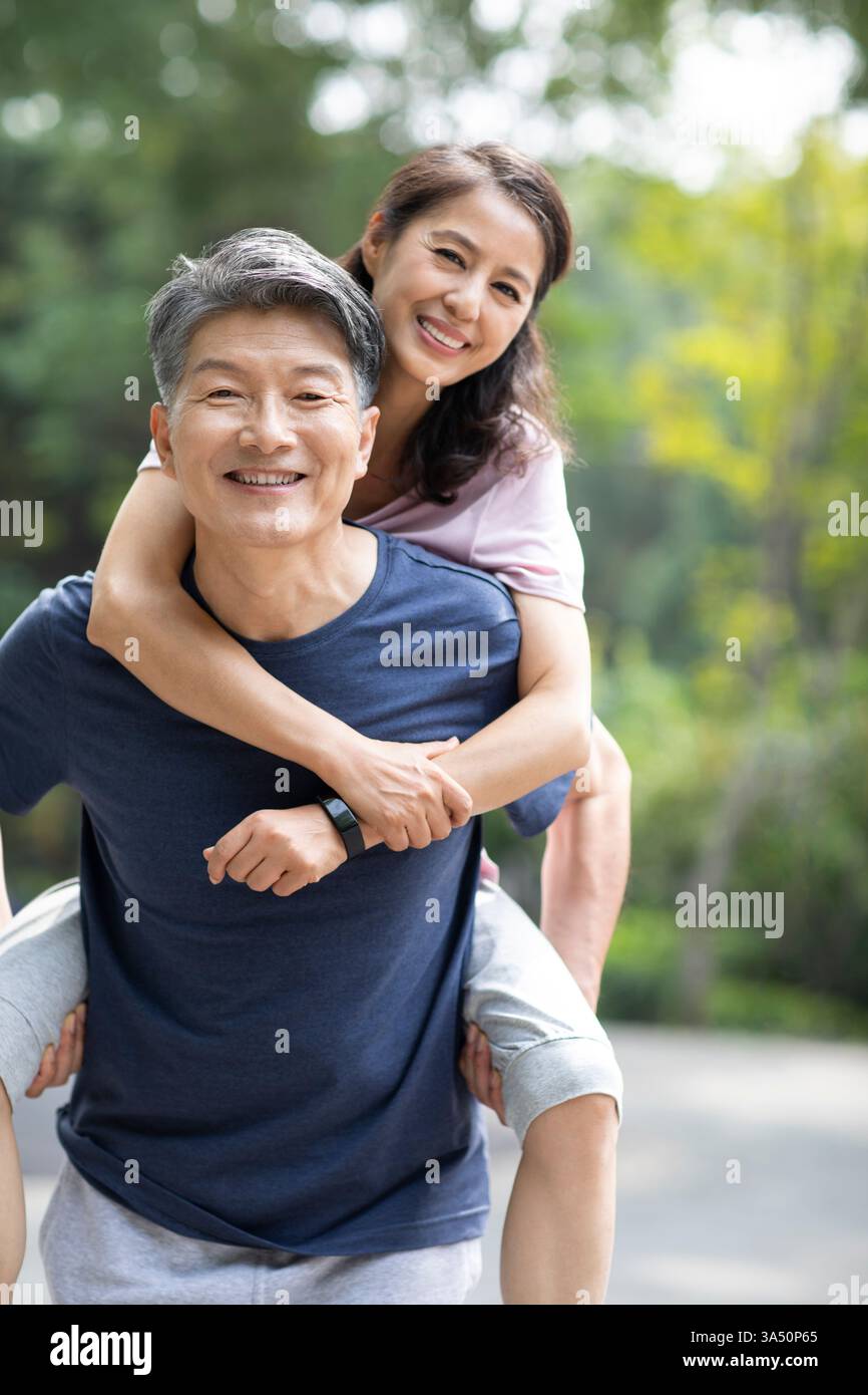 Smiling senior Chinese man giving piggy back ride to her middle-aged Chinese wife while standing ...