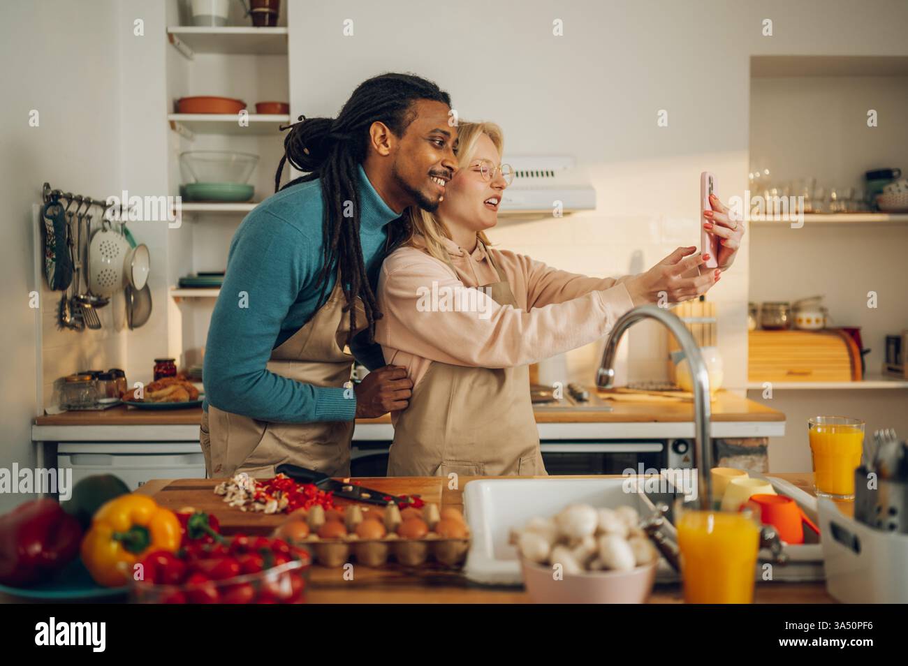 The multiracial partners are standing in a kitchen at home and taking ...