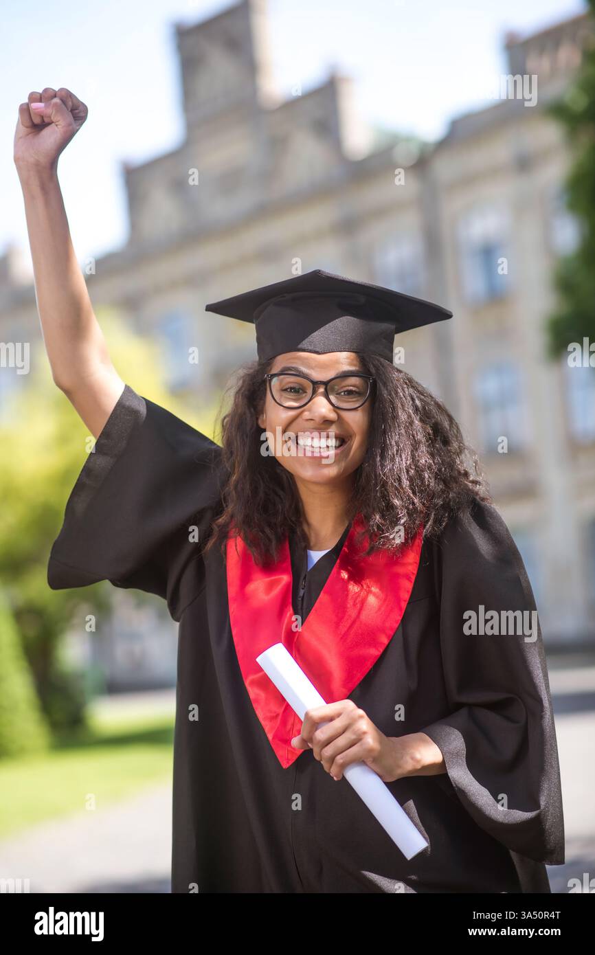 Cheerful African American female in graduation gown raising closed fist ...