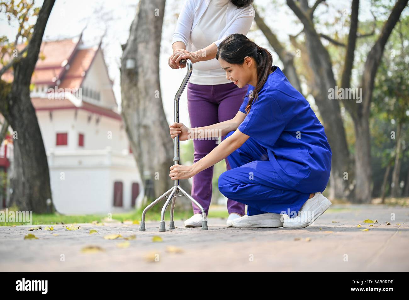 Smiling Asian female caregiver in blue uniform crouching and helping ...