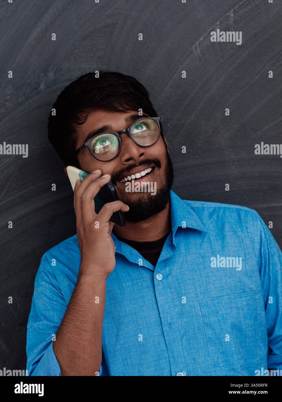 Indian smiling young student in blue shirt and glasses using smartphone ...