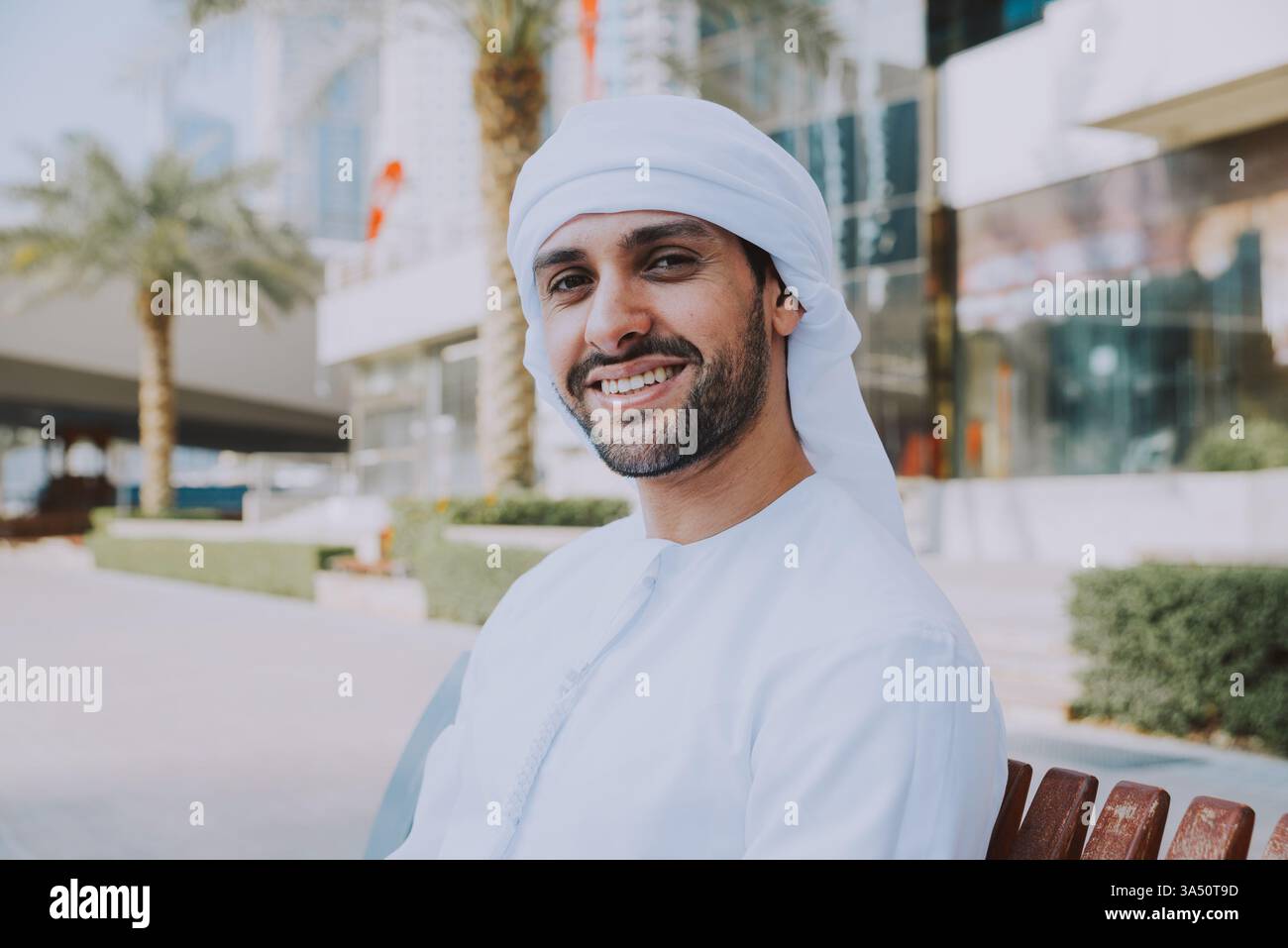 Smiling Middle Eastern businessman sitting on bench near city buildings ...