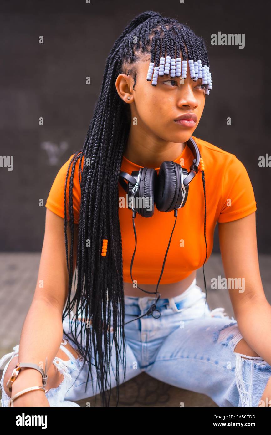 Native American woman with braids and headphones on neck looking away sitting on bench in city ...